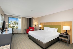 Quarto de hotel com uma cama grande, uma escrivaninha com computador, uma cadeira vermelha e vista para a cidade.