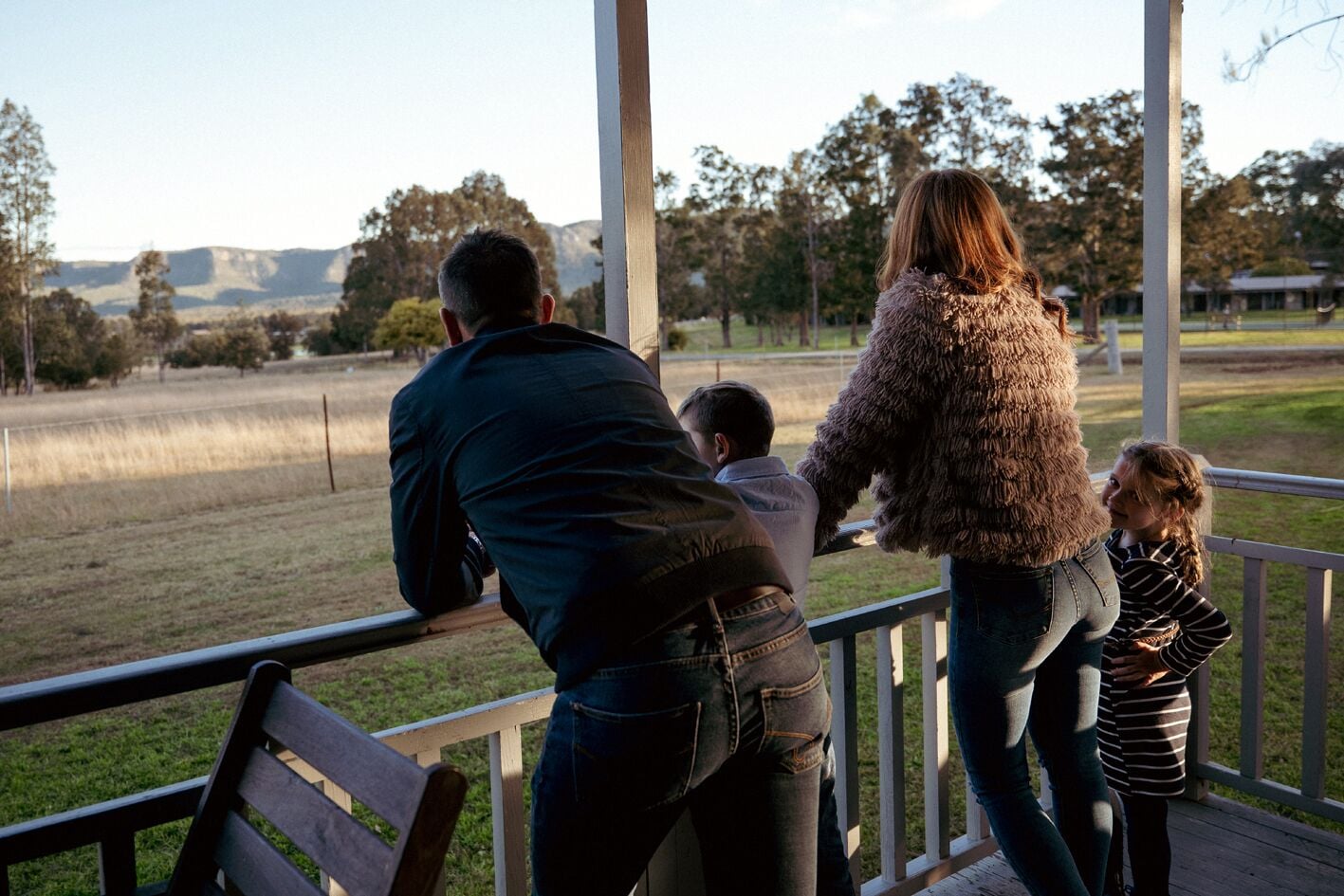 Uma família numa varanda com vista para um campo gramado, árvores e colinas ao longe.