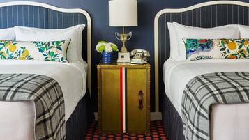 Quarto de hotel com duas camas, cada uma com travesseiro estampado com flores, uma pequena mesa de cabeceira com luminária e um telefone antigo.