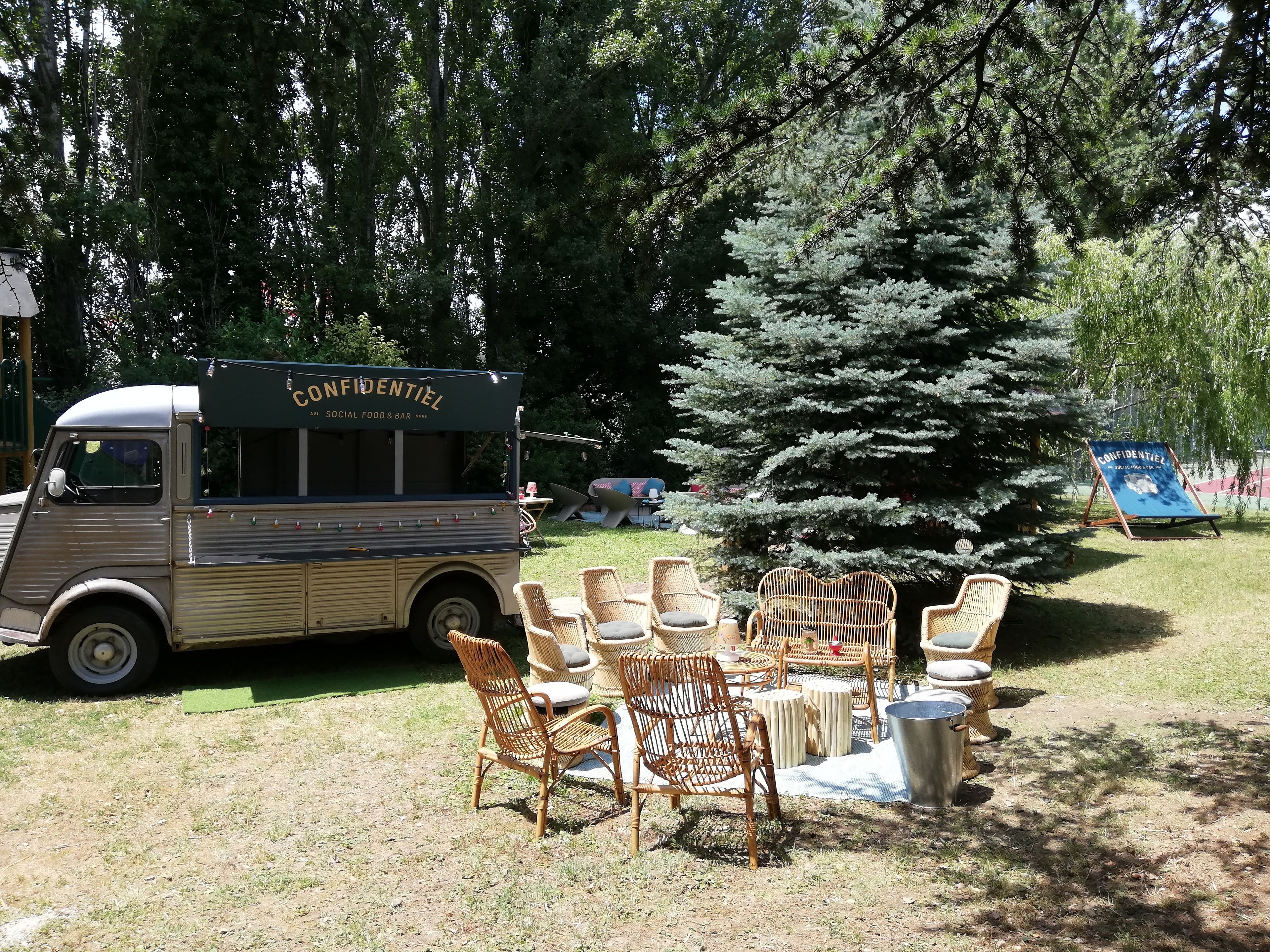 Um food truck antigo chamado 'Confidentiel', com móveis de vime e uma grande árvore de Natal em um parque.