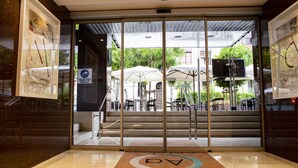 Entrada de hotel com porta de vidro, rampa e área externa com mesas e guarda-sóis.