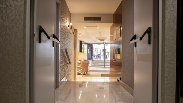 Corredor moderno de hotel com piso polido, área de recepção com balcão e cadeiras, e vista para a área de estar externa.