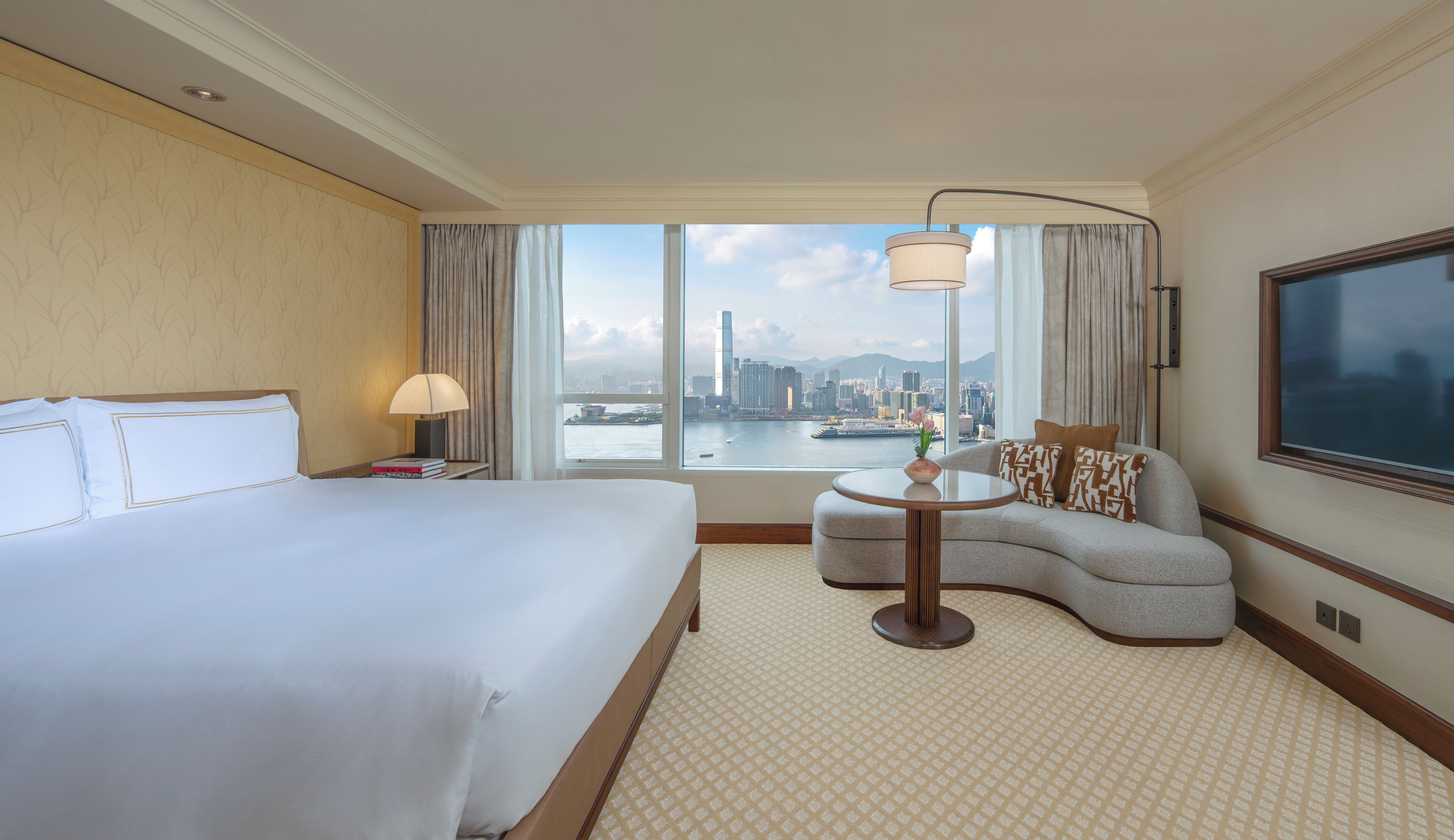 Quarto de hotel com uma cama grande, um sofá, uma mesinha e uma televisão. Há uma janela com vista para a cidade.