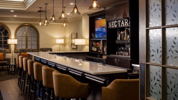 Uma área de bar com um balcão comprido, vários bancos altos, luminárias pendentes e um bar abastecido com garrafas.