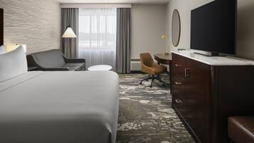 Quarto de hotel com uma cama grande, uma escrivaninha, uma cadeira, uma televisão e piso coberto por carpete.