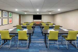 Uma sala de conferências com mesas, cadeiras, tela de projetor e quadros nas paredes.