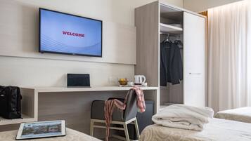 Quarto de hotel com TV exibindo 'WELCOME', escrivaninha com laptop, cadeira, cama e guarda-roupa com roupas penduradas.