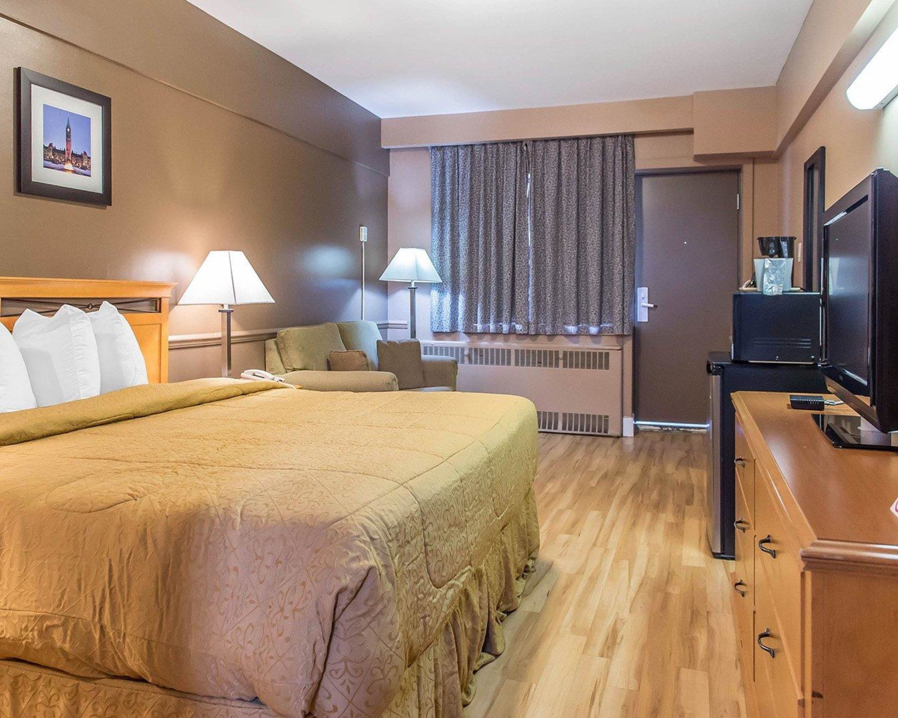 Quarto de hotel com uma cama, duas luminárias de cabeceira, uma televisão, uma janela com cortinas e um quadro na parede.