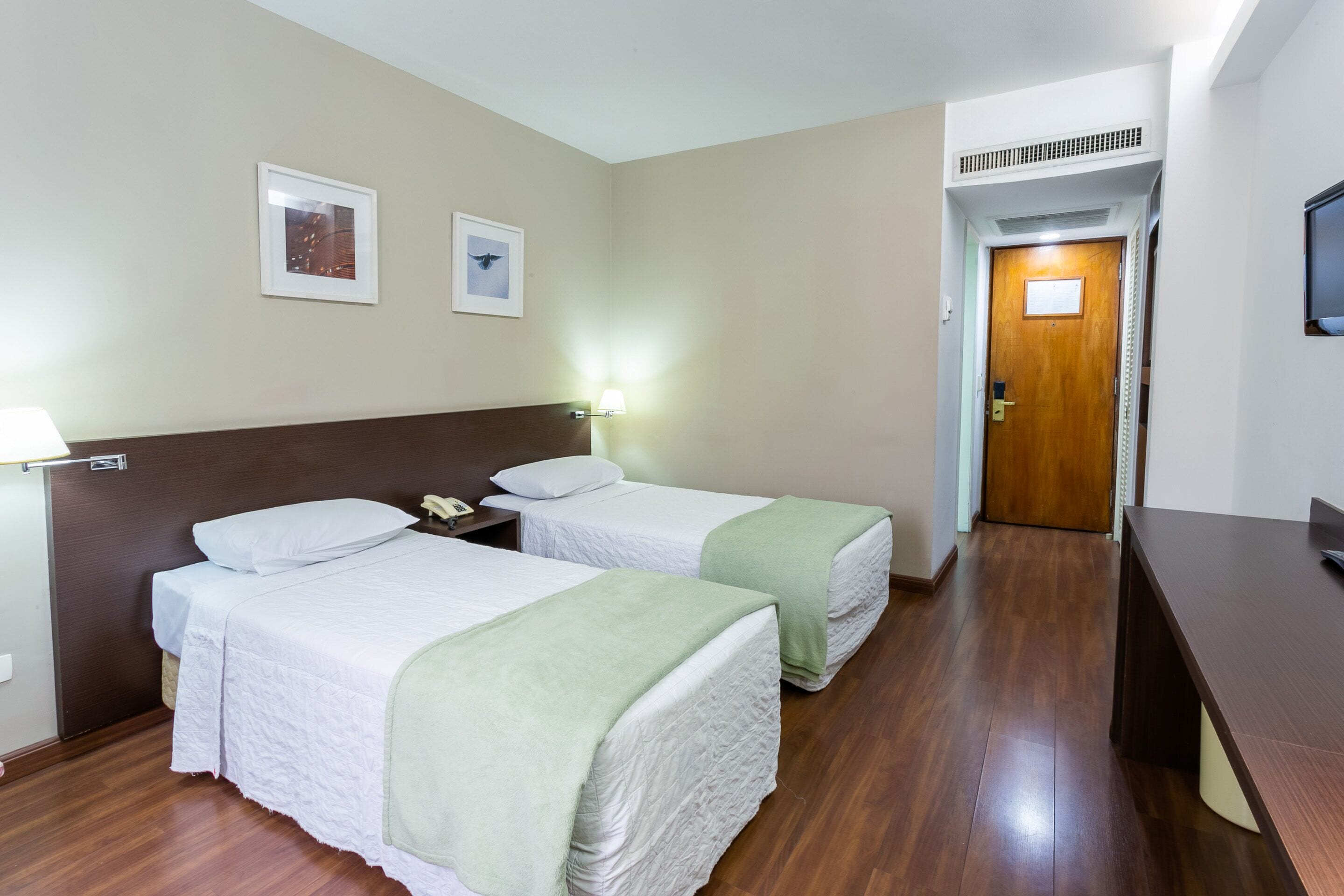 Quarto de hotel com duas camas, uma mesa, uma televisão e uma porta que dá para outro espaço.