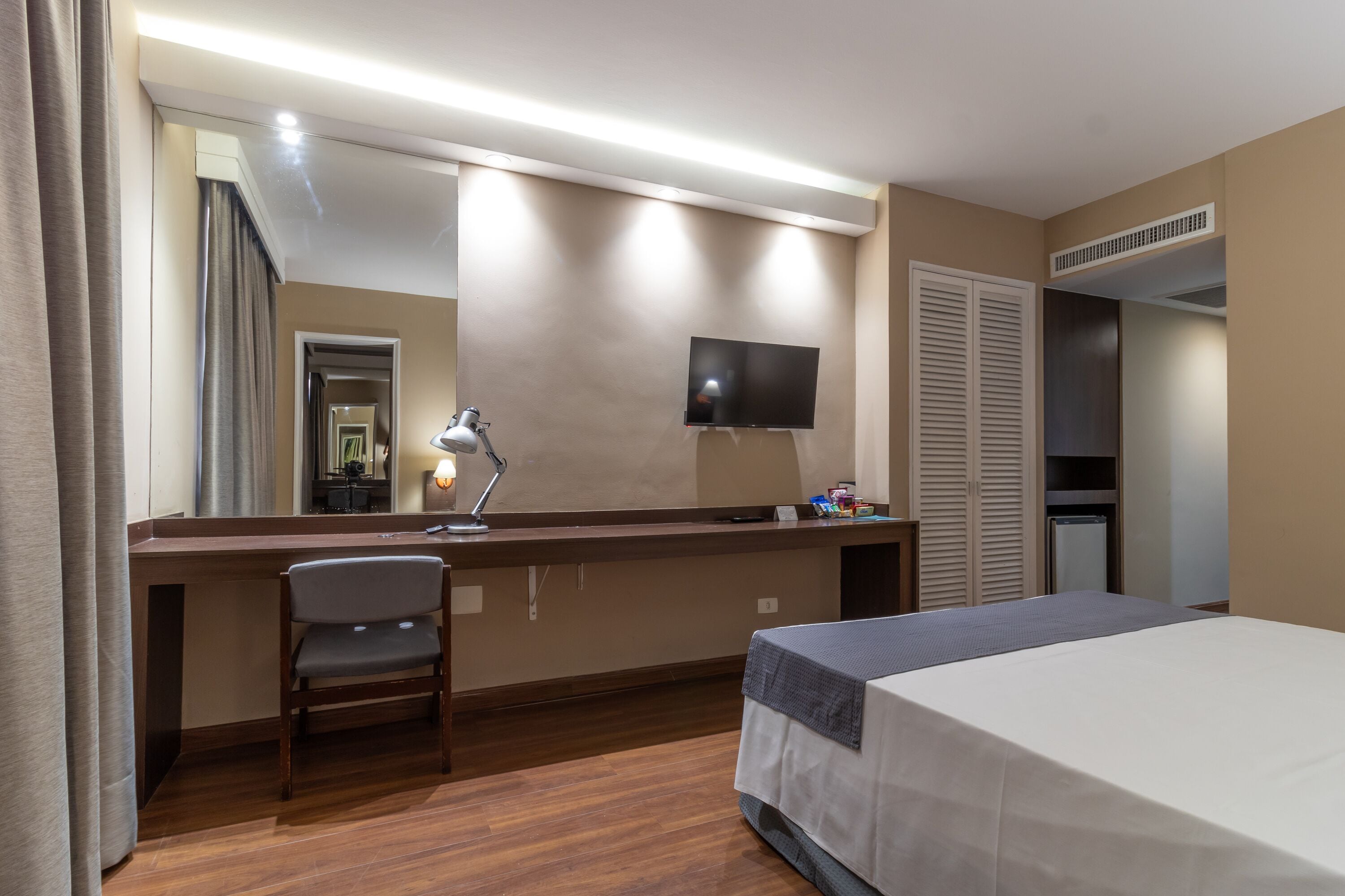 Quarto de hotel com cama, escrivaninha, cadeira, TV e mini-geladeira.