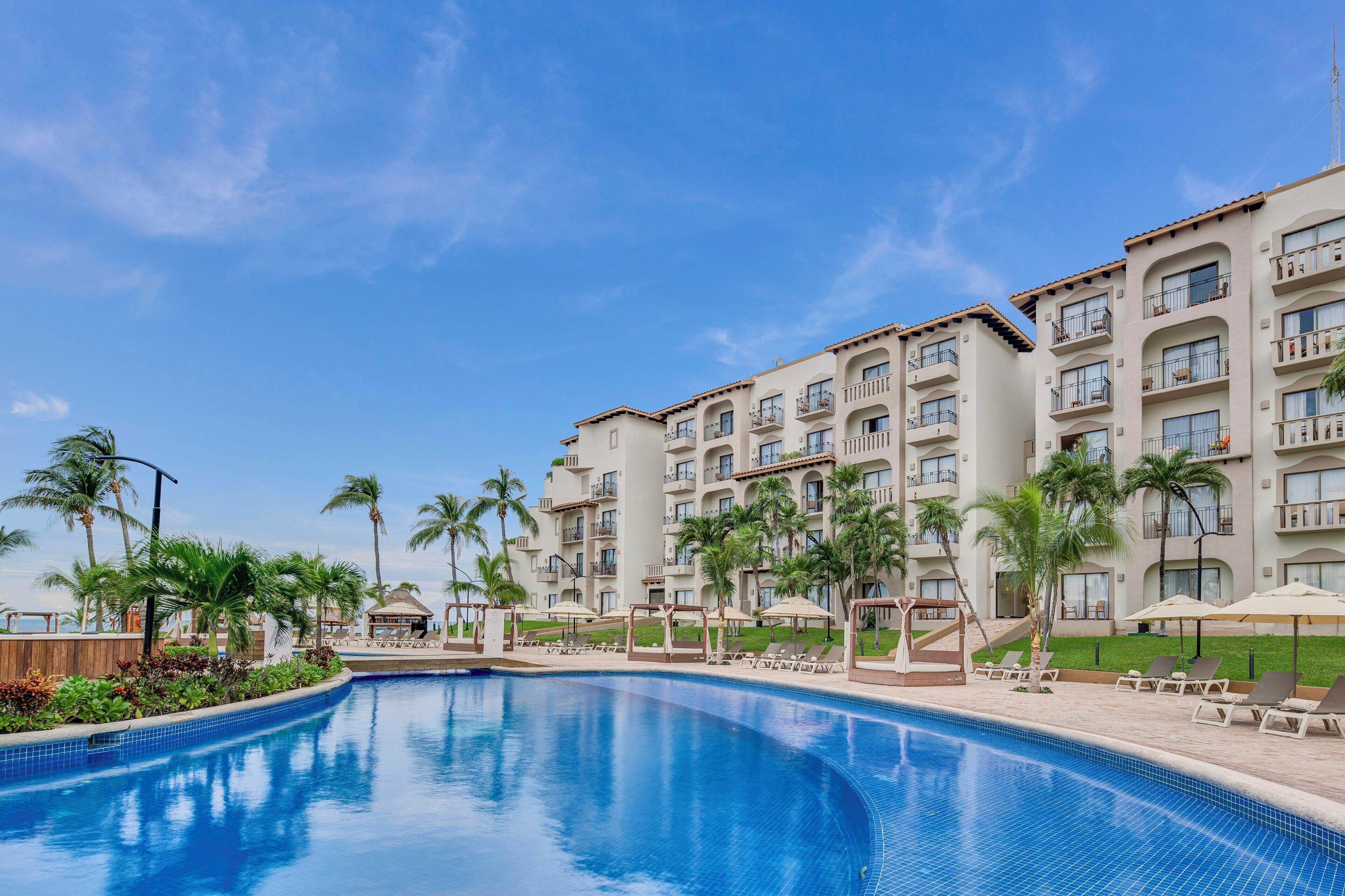 Um hotel com piscina, palmeiras e um céu azul límpido.