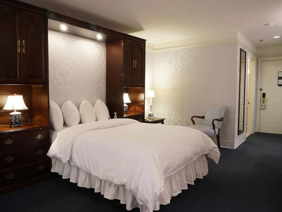 Quarto de hotel com uma cama grande, duas mesas de cabeceira com luminárias, uma cadeira e uma porta.