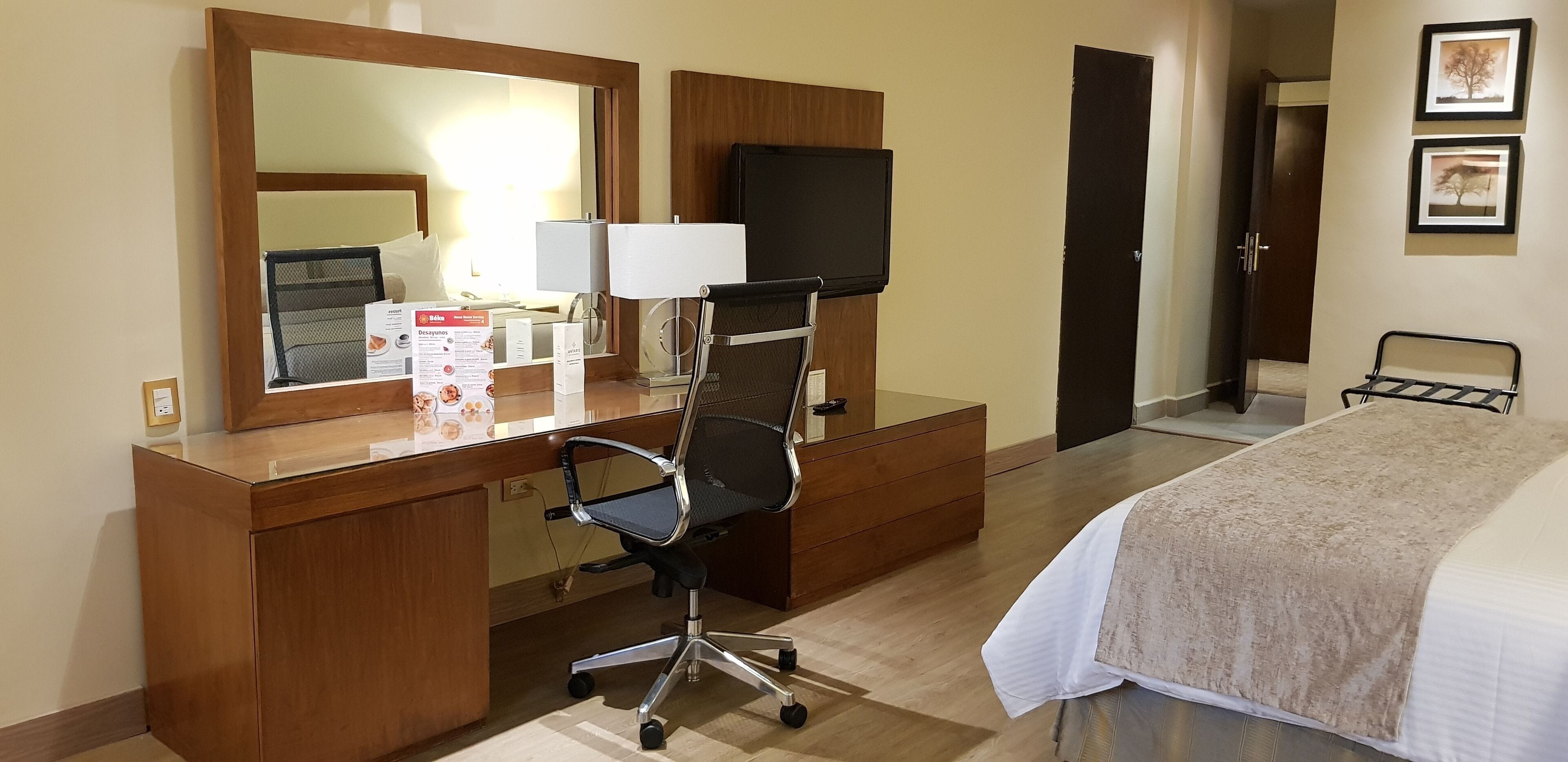 Quarto de hotel com escrivaninha de madeira, uma cadeira, uma cama com manta bege, televisão e porta que dá para outro cômodo.