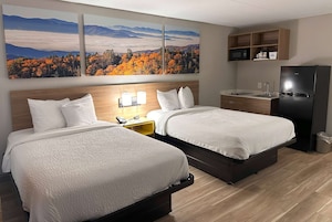 Quarto de hotel com duas camas, cabeceira de madeira, escrivaninha pequena com luminária, micro-ondas e geladeira.