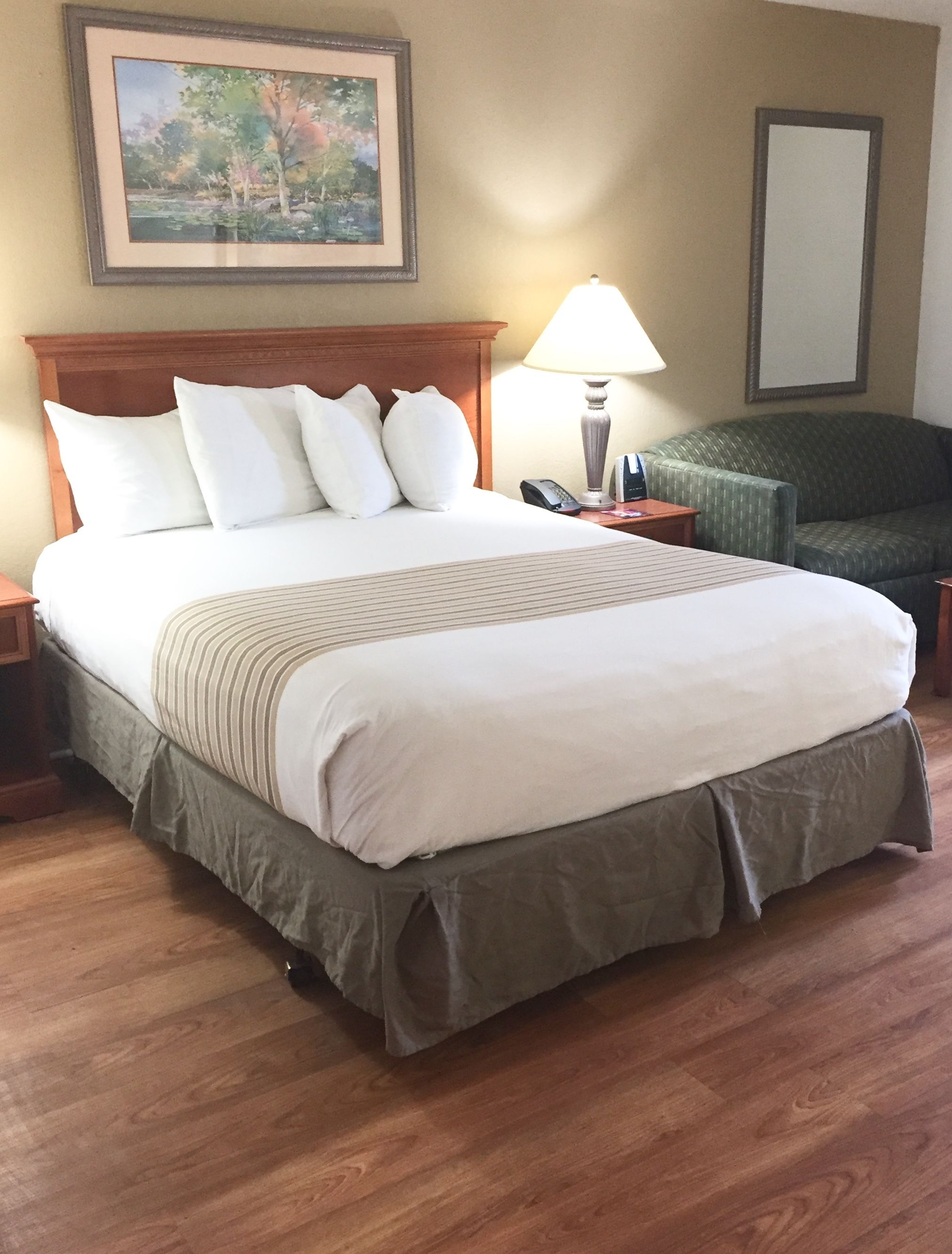 Quarto de hotel com uma cama grande, uma mesa de cabeceira com abajur, uma poltrona verde e um quadro emoldurado na parede.