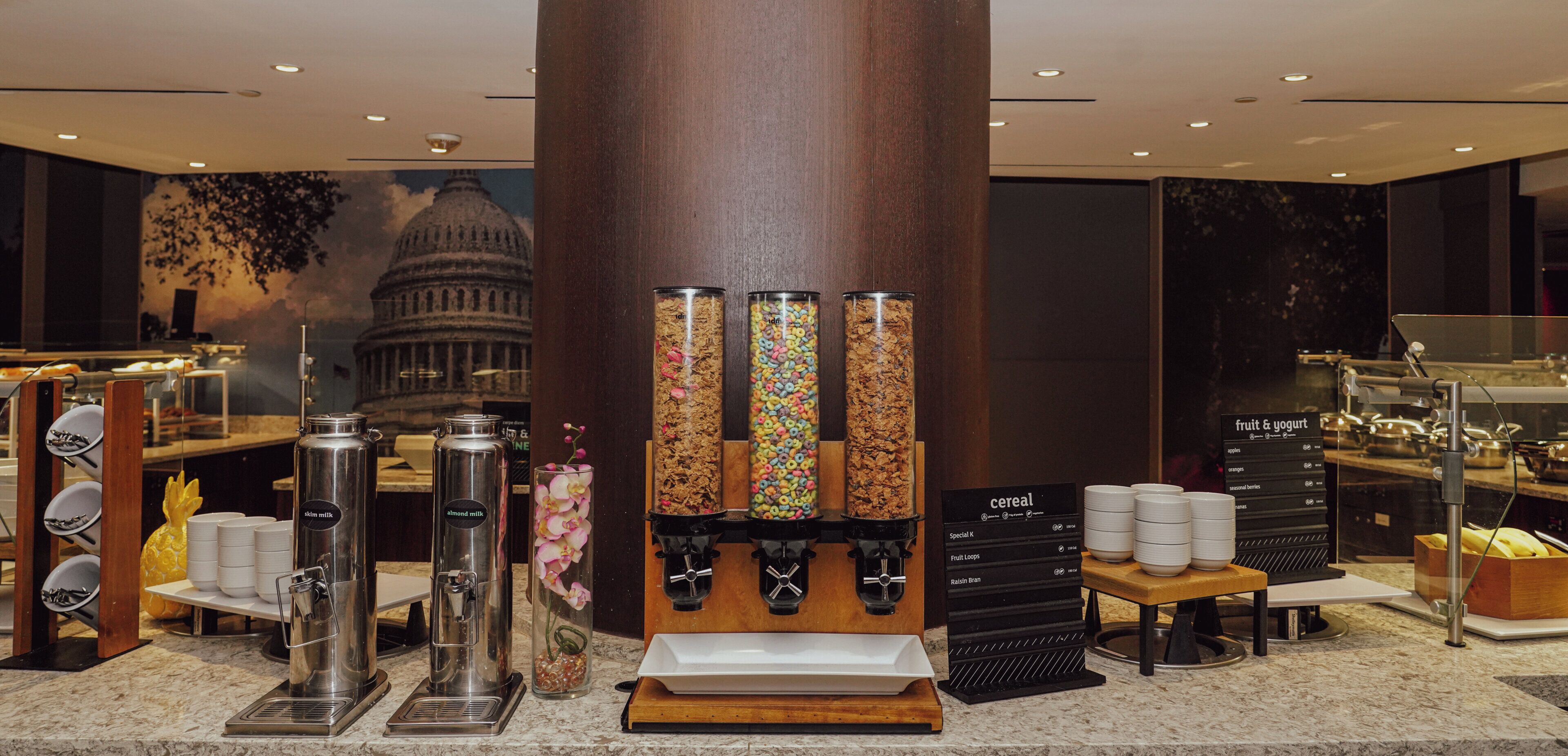 Um balcão de café da manhã com máquina de café, distribuidores de cereais e uma estação de frutas e iogurte.