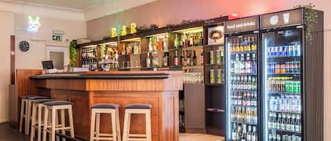 Uma área de bar com banquetas, balcão e duas geladeiras repletas de bebidas.