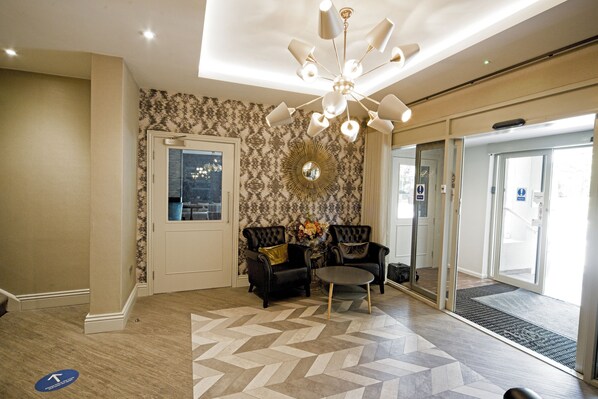 Um hall com parede estampada, um candelabro, duas poltronas e uma porta que dá para o exterior.