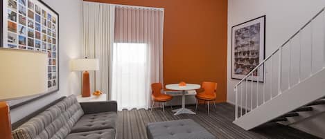 Quarto de hotel com sofá cinzento, mesa redonda pequena com duas cadeiras, fotografia grande emoldurada na parede e uma escada.