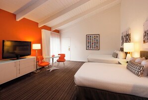 Quarto de hotel com televisão, duas camas, escrivaninha com cadeira e fotografia emoldurada na parede.