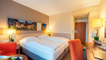 Quarto de hotel com cama, secretária com laptop, cadeira, mesa com fruta e duas luminárias fixadas na parede.