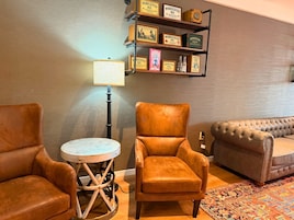 Quarto com poltrona de couro marrom, mesa redonda, sofá com capitonê e estante com objetos antigos.