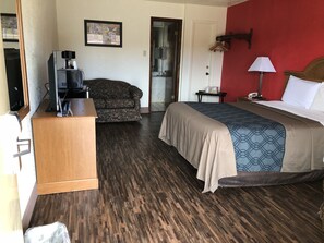 Quarto de hotel com cama, escrivaninha com computador, sofá, parede de destaque vermelha e porta para um banheiro.