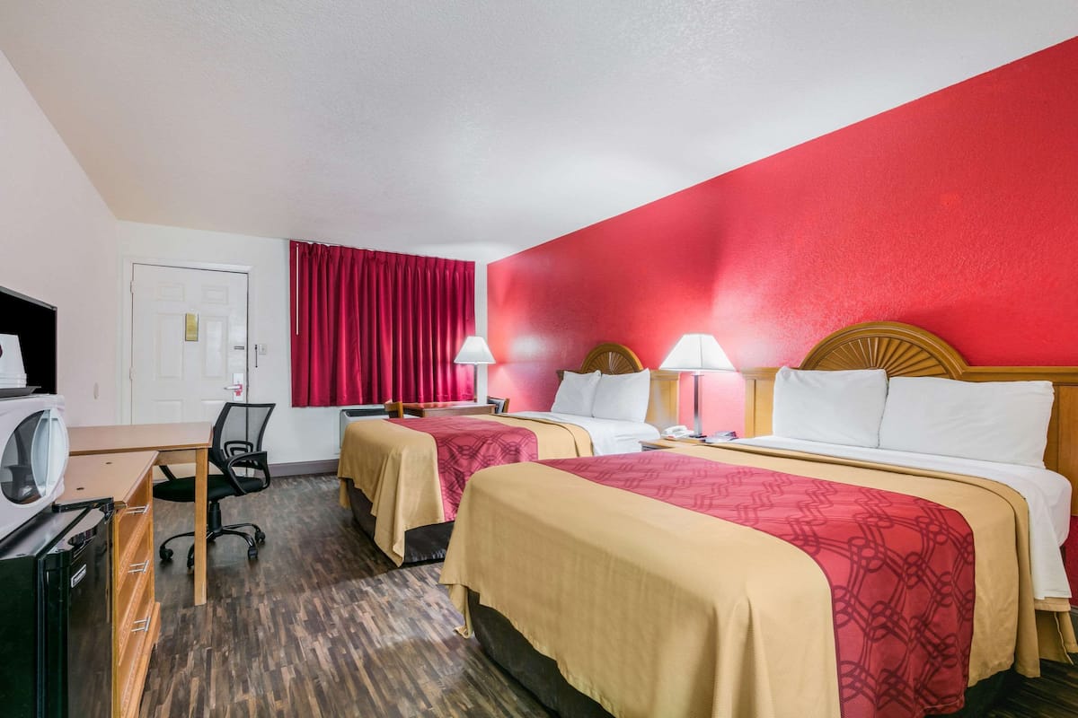 Quarto de hotel com duas camas, uma escrivaninha com cadeira, televisão e cortina vermelha.