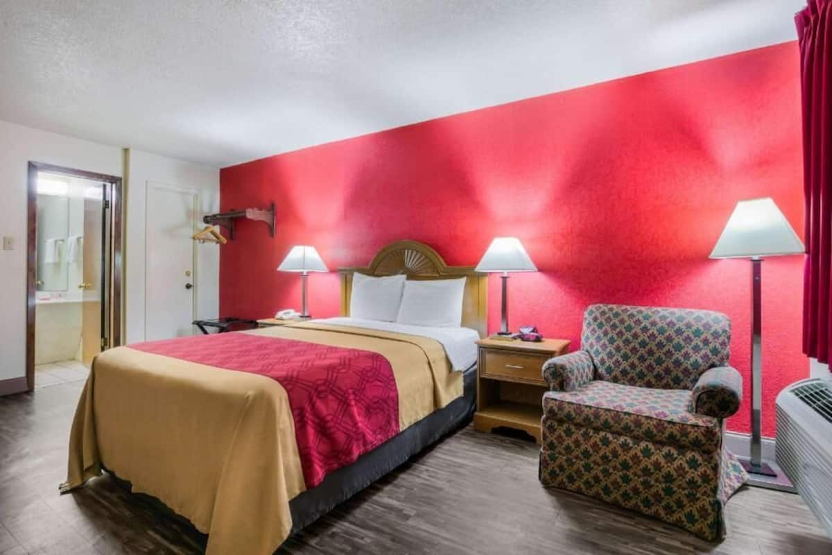 Quarto de hotel com cama, duas luminárias de cabeceira, uma cadeira, mesa de cabeceira e uma parede de destaque vermelha.