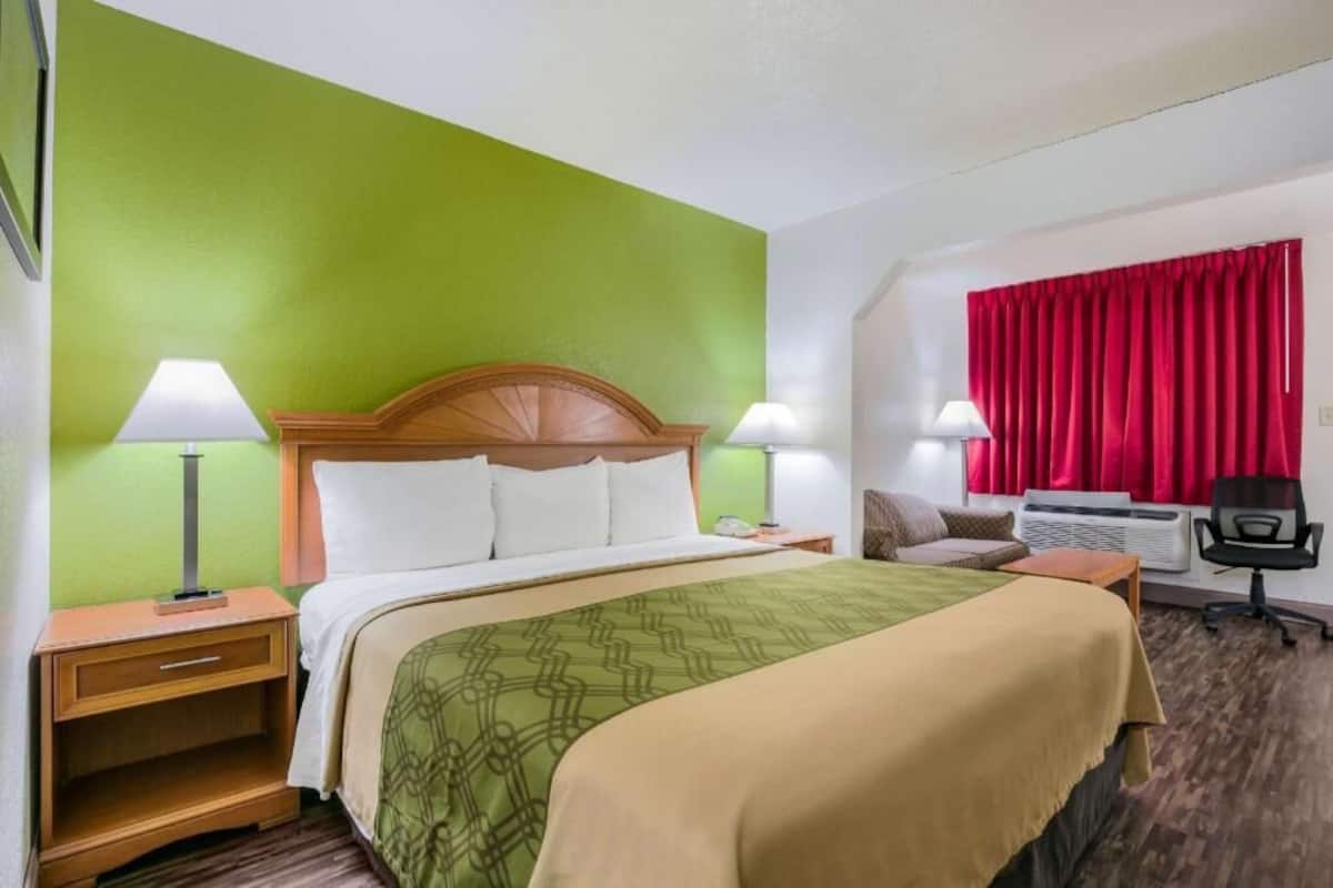 Quarto de hotel com uma cama, duas mesas de cabeceira, uma escrivaninha, uma cadeira e uma janela com cortinas vermelhas.