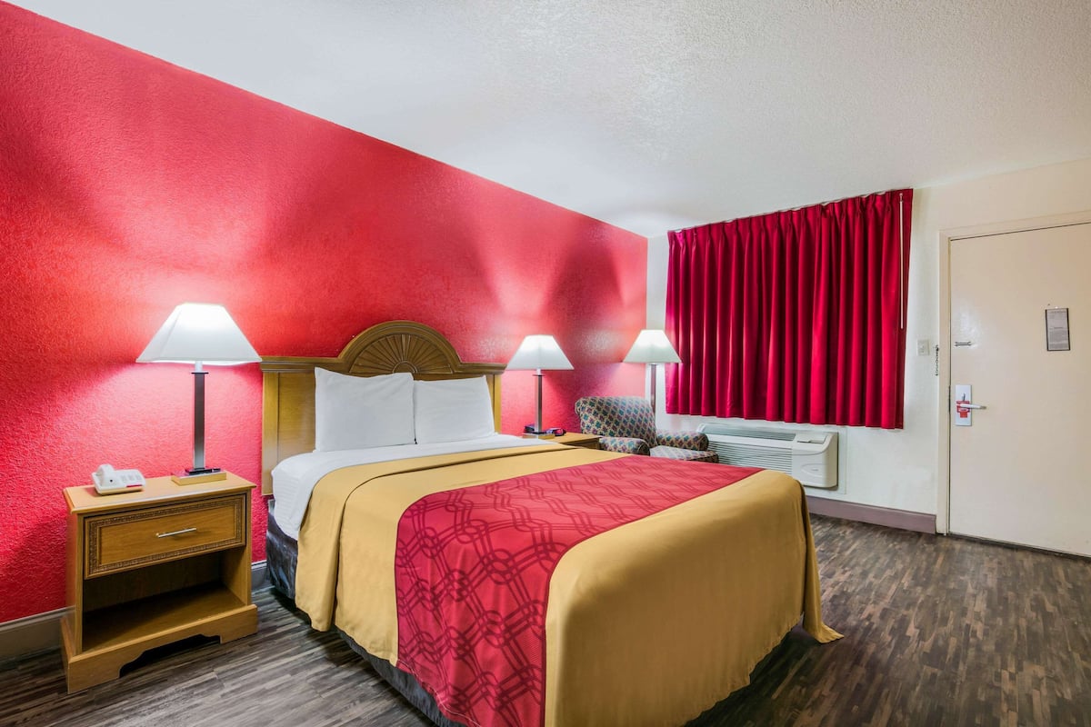 Quarto de hotel com parede de destaque vermelha, cama com colcha vermelha e amarela, duas luminárias de cabeceira, mesa de cabeceira com telefone e janela com cortinas vermelhas.
