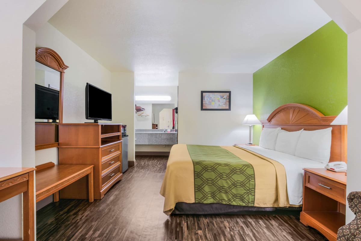 Quarto de hotel com cama, escrivaninha, cadeira, televisão e banheiro visível ao fundo.
