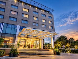 Entrada de hotel com fachada de vidro, degraus levando à entrada e área coberta com teto.
