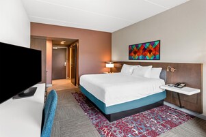 Quarto de hotel com cama, televisão, escrivaninha, abajur e uma obra de arte colorida na parede.