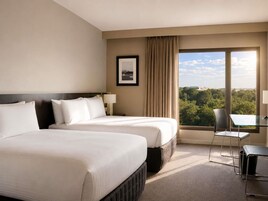 Quarto de hotel com duas camas, uma escrivaninha, uma cadeira, janela com vista e um quadro na parede.