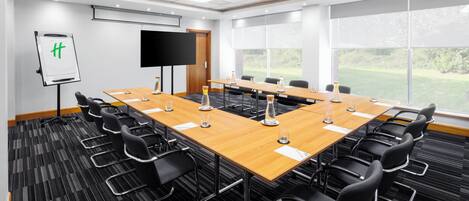 Uma sala de conferência com uma mesa comprida, cadeiras pretas, um quadro branco e uma janela grande com vista para o verde.