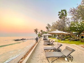 Resort à beira-mar com espreguiçadeiras e guarda-sóis, um caminho de pedrinhas e palmeiras.
