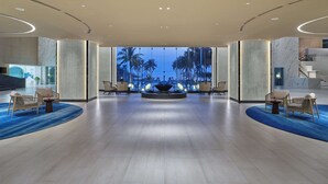 Um hall amplo com área de estar central, grandes janelas com vista para palmeiras e o mar, e um design arquitetônico moderno.
