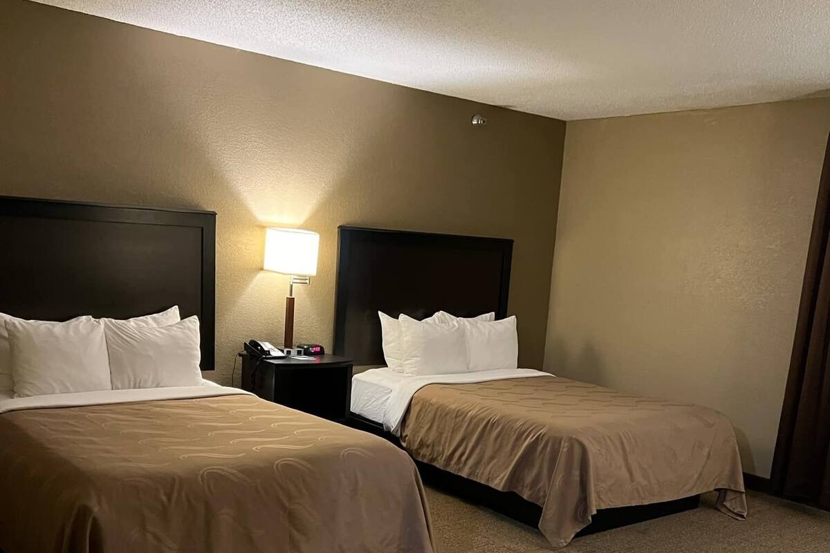 Quarto de hotel com duas camas, uma mesinha de cabeceira e uma luminária.