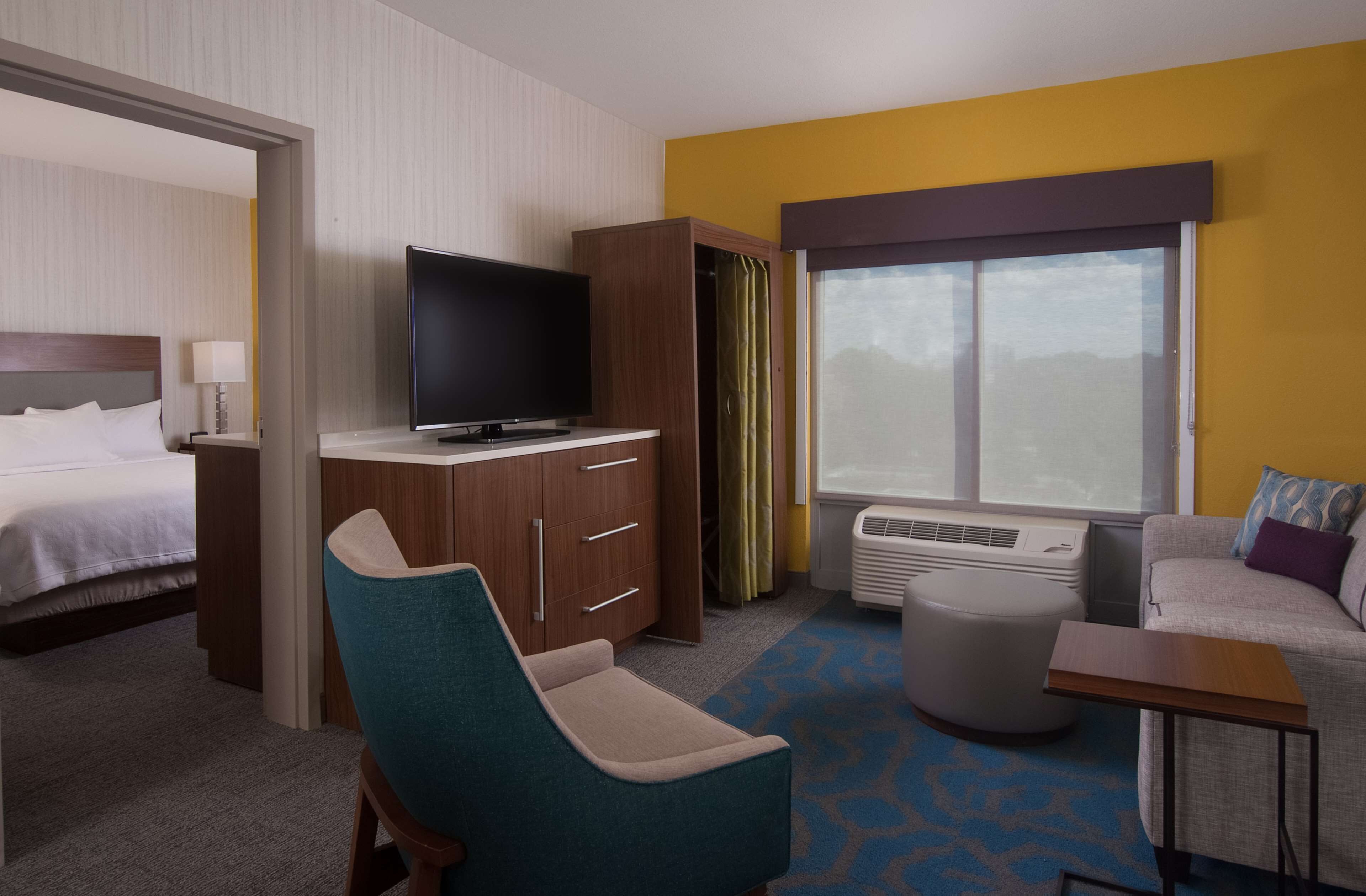 Quarto de hotel com cama, televisão, escrivaninha, cadeira, sofá e janela com cortinas.