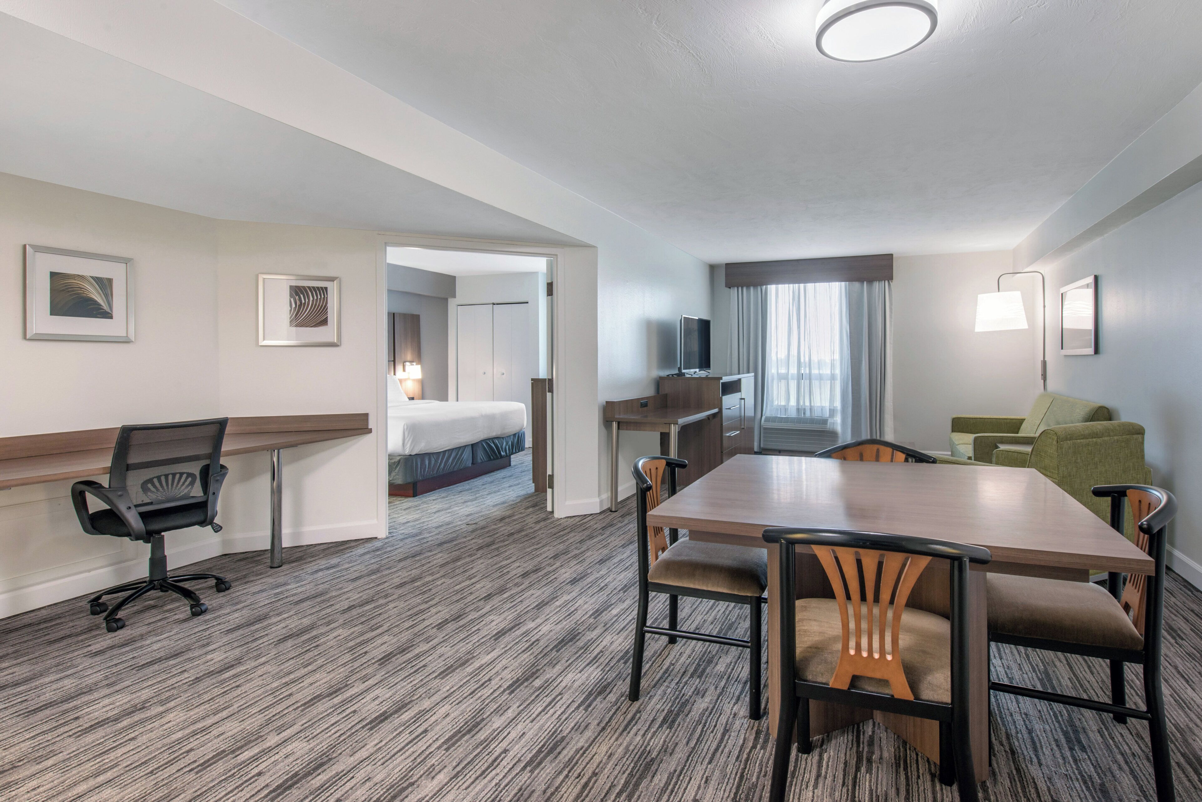 Quarto de hotel com cama, escrivaninha, mesa de jantar com cadeiras, televisão e janela com cortinas.