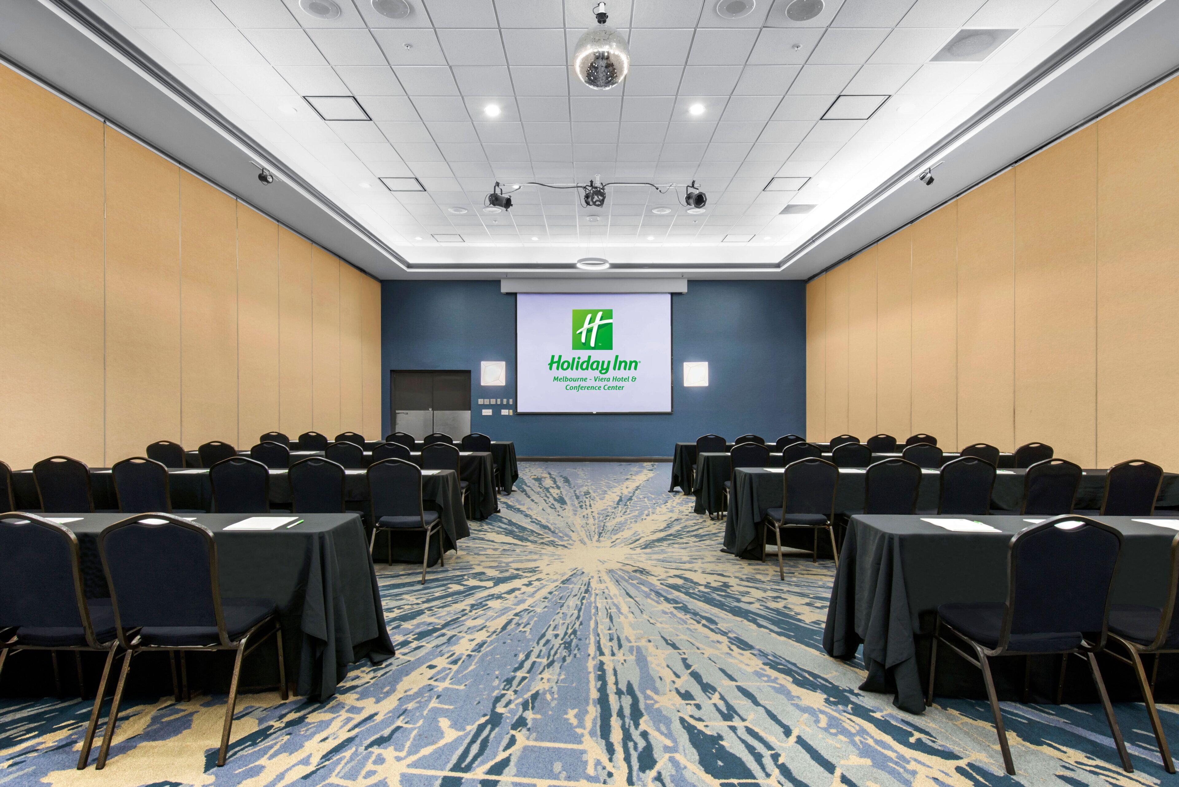 Uma sala de conferências com fileiras de mesas e cadeiras, uma tela grande exibindo o logotipo do Holiday Inn, e piso coberto de carpete.