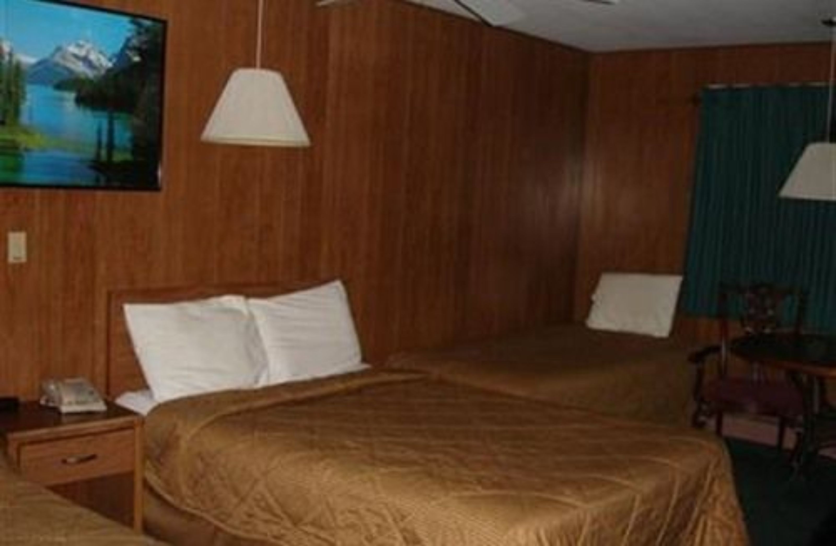 Quarto de hotel com painéis de madeira, cama com colcha marrom, duas mesas de cabeceira, uma cadeira e um quadro com paisagem de montanhas.