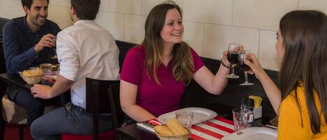 Um grupo de pessoas jantando em um restaurante com toalha quadriculada vermelha e branca, uma cesta de pão e taças de vinho na mesa.