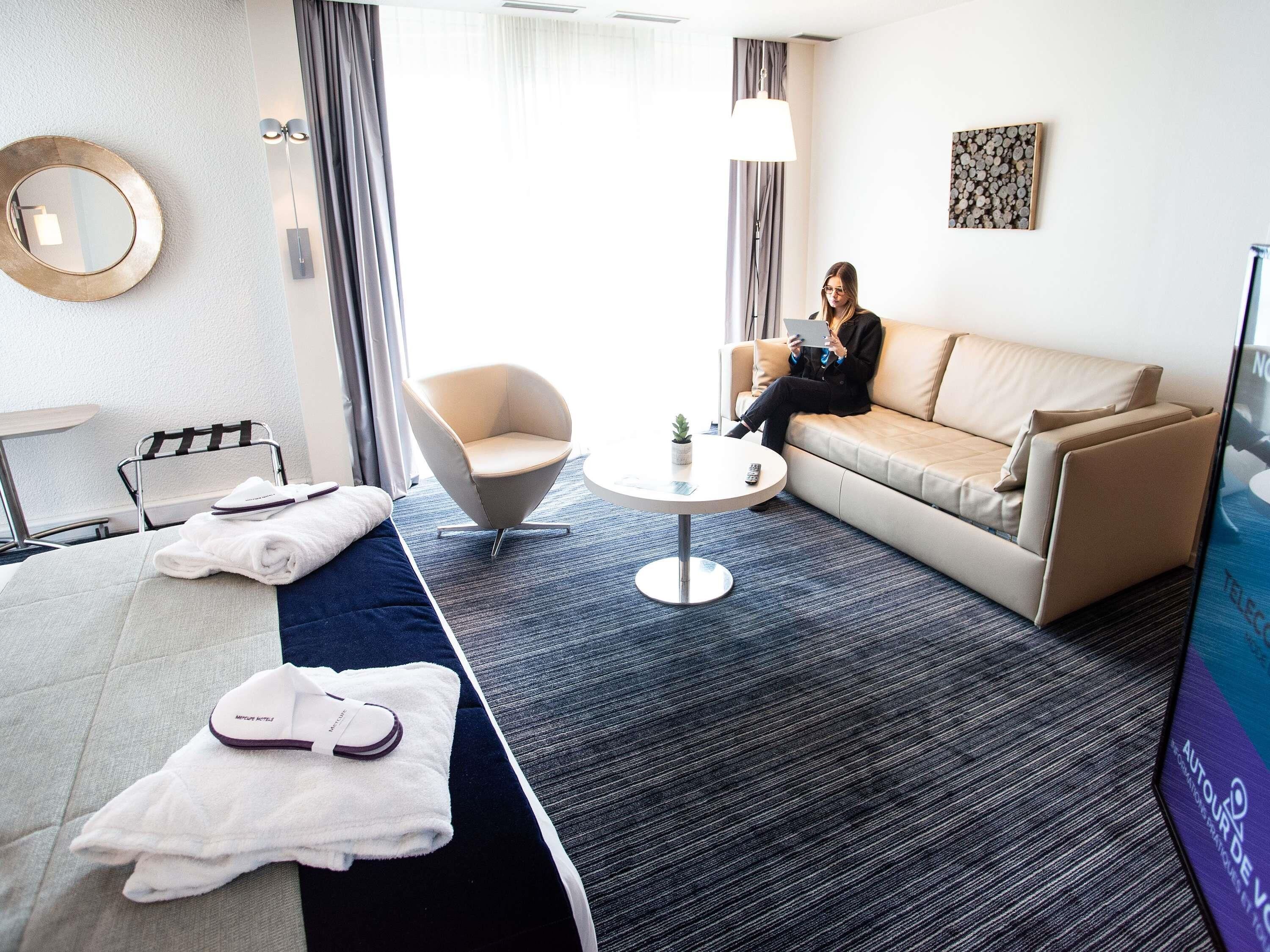 Quarto de hotel com cama, área de estar com sofá e poltrona, mesa redonda pequena e televisão.