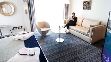 Quarto de hotel com cama, área de estar com sofá e poltrona, mesa redonda pequena e televisão.