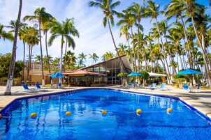 Piscina com água azul cristalina, rodeada por espreguiçadeiras e guarda-sóis, com palmeiras e um edifício ao fundo.