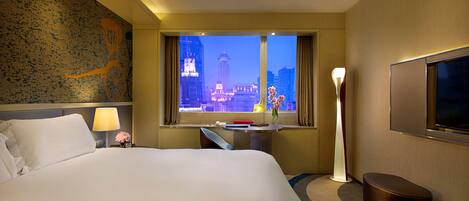 Quarto de hotel com cama, escrivaninha, televisão, janela com vista para a cidade e luminária.