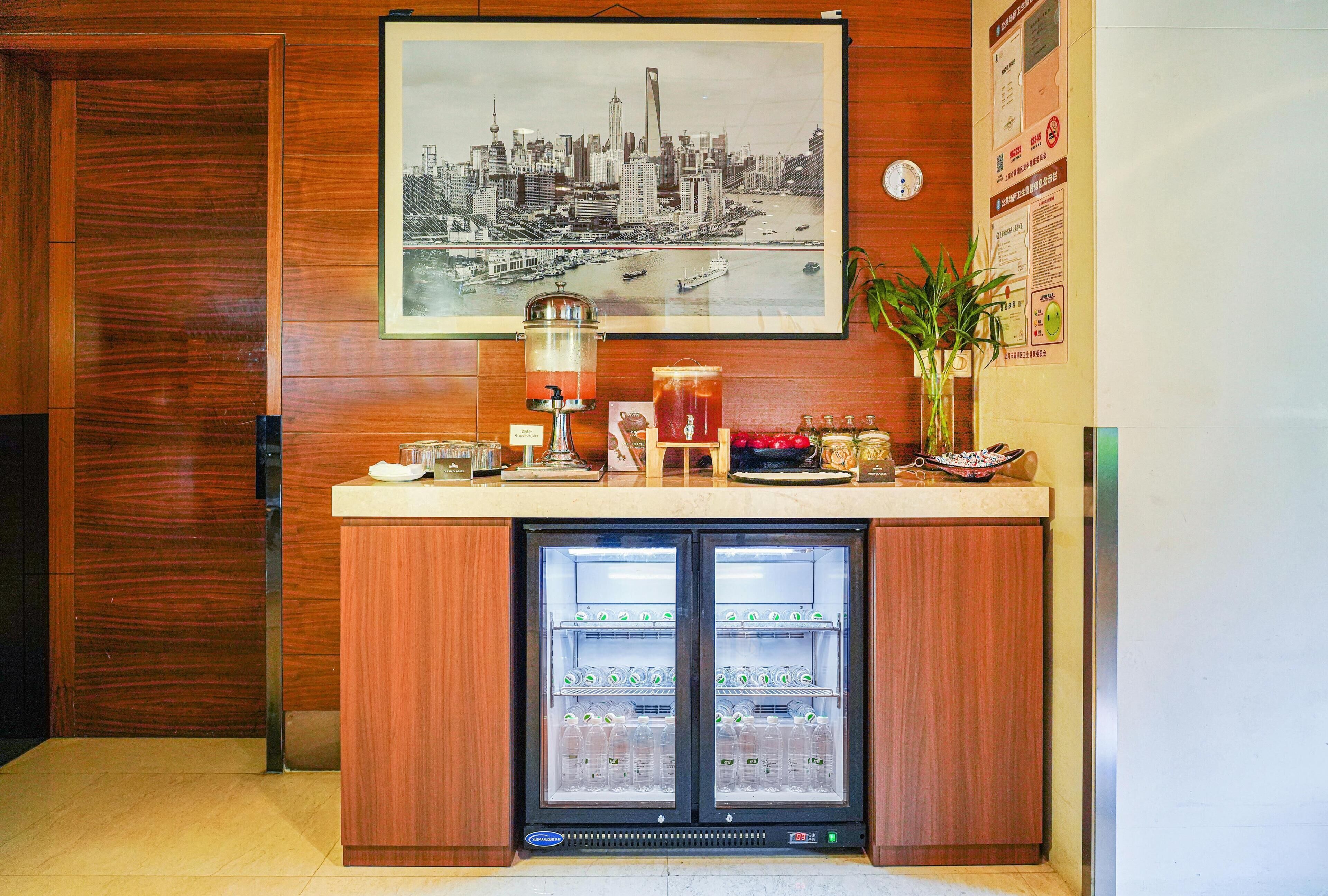 Uma área de recepção de hotel com painel de parede de madeira, uma fotografia emoldurada da paisagem urbana, um dispensador de bebidas e uma geladeira abastecida com água engarrafada.