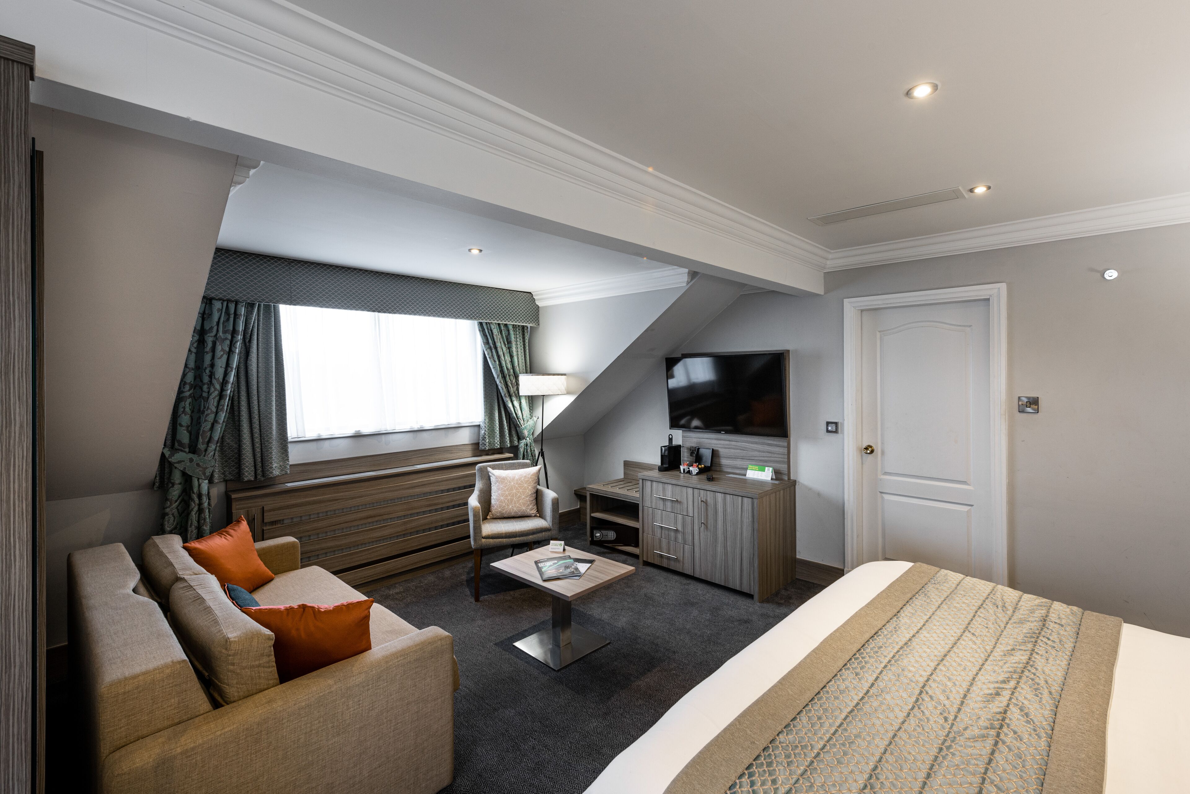 Quarto de hotel moderno com sofá, poltrona e cama. Há uma televisão e uma pequena escrivaninha com abajur.